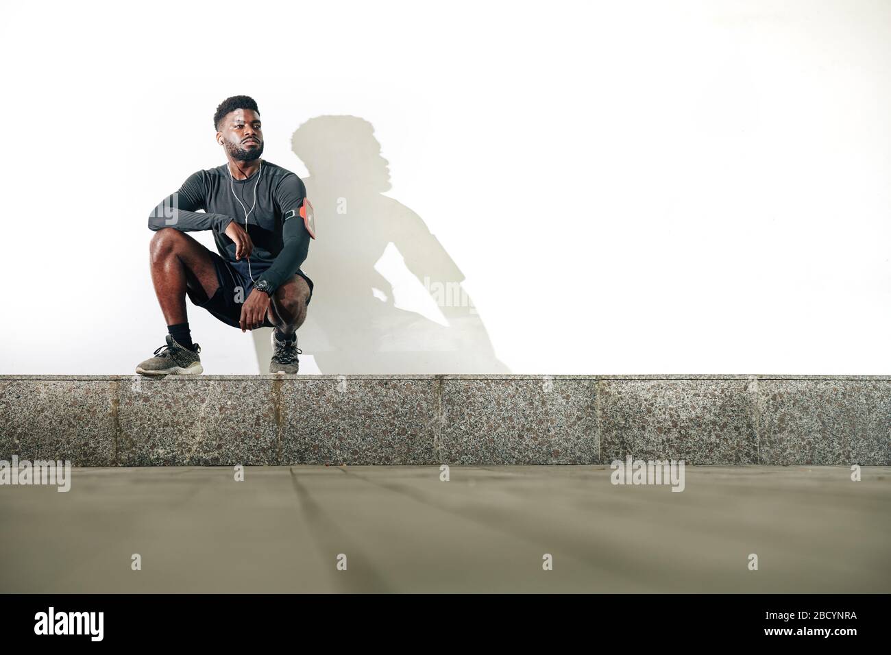 Serious confident young Black sportsman sitting on haunches on kerb ...