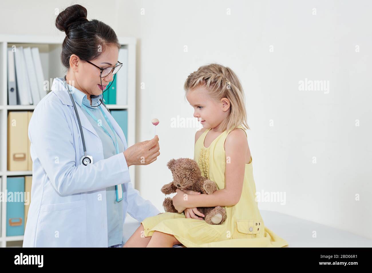 Smiling pediatrician giving lollipop to little girl trying to ease her ...