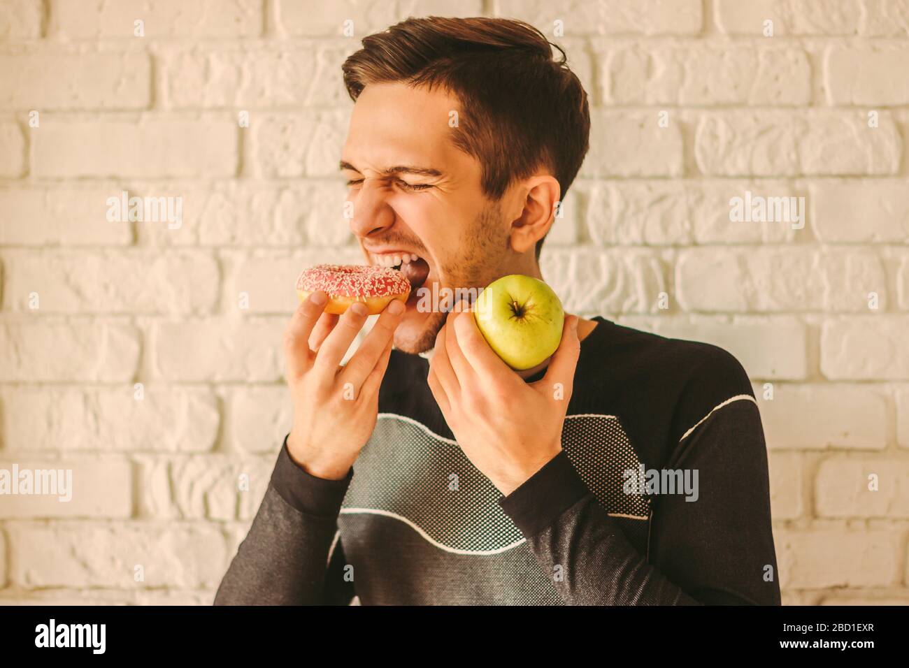Young attractive sports man eating donut cake and holding apple in hand ...