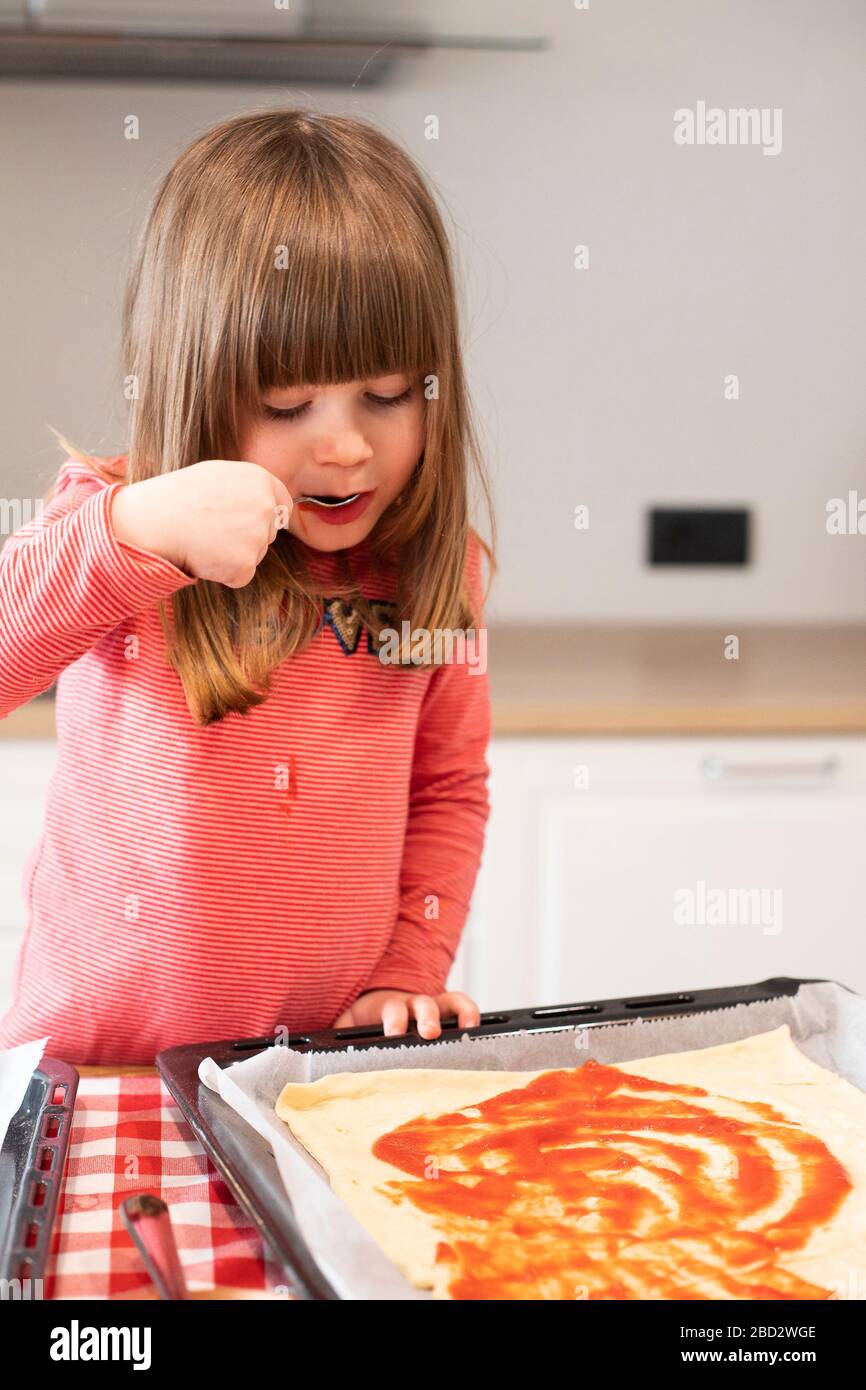 Child girl in a kitchen tasting some tomato sauce from raw pizza before ...