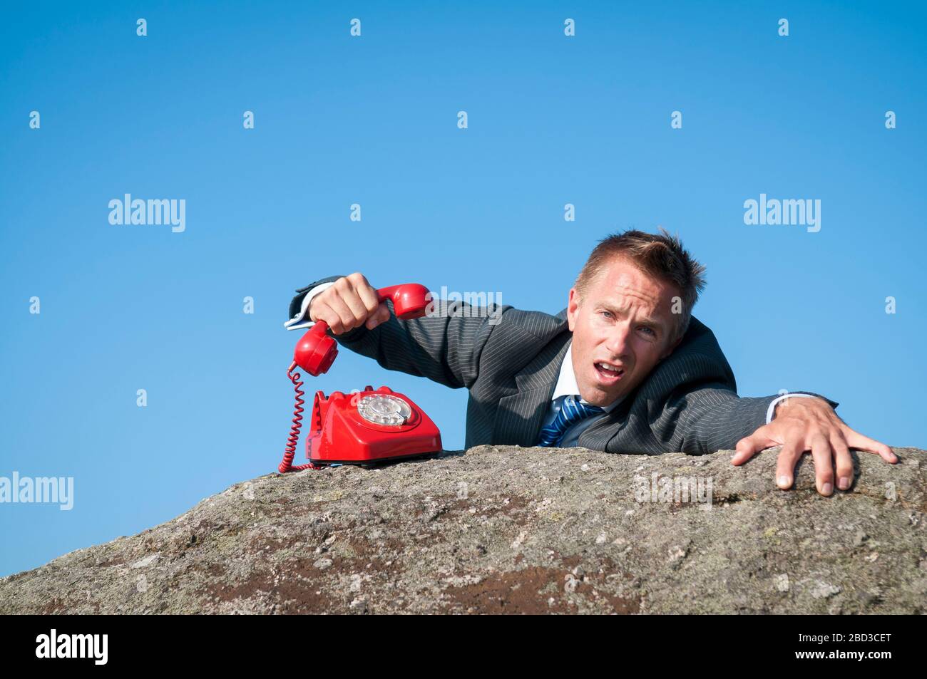 Businessman dangling off a rock cliff reaching to make a desperate call ...