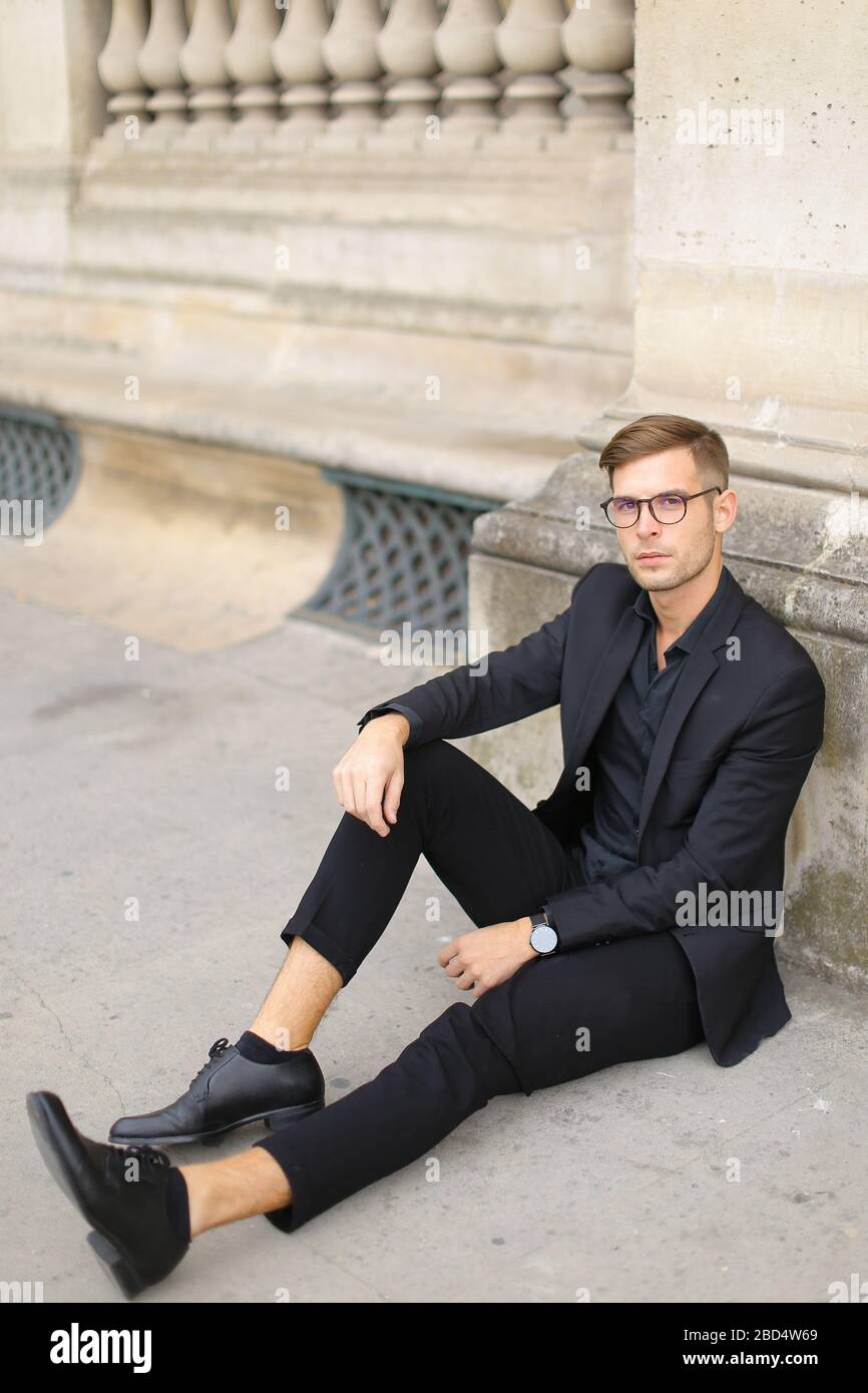 Young european man sitting on sidewalk ground and wearing black suit ...
