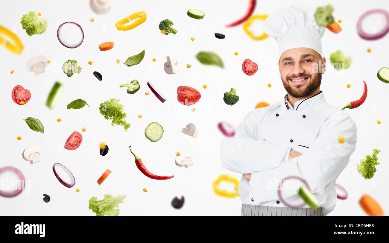Healthier cooking. Smiling chef in uniform and flying veggies on white ...