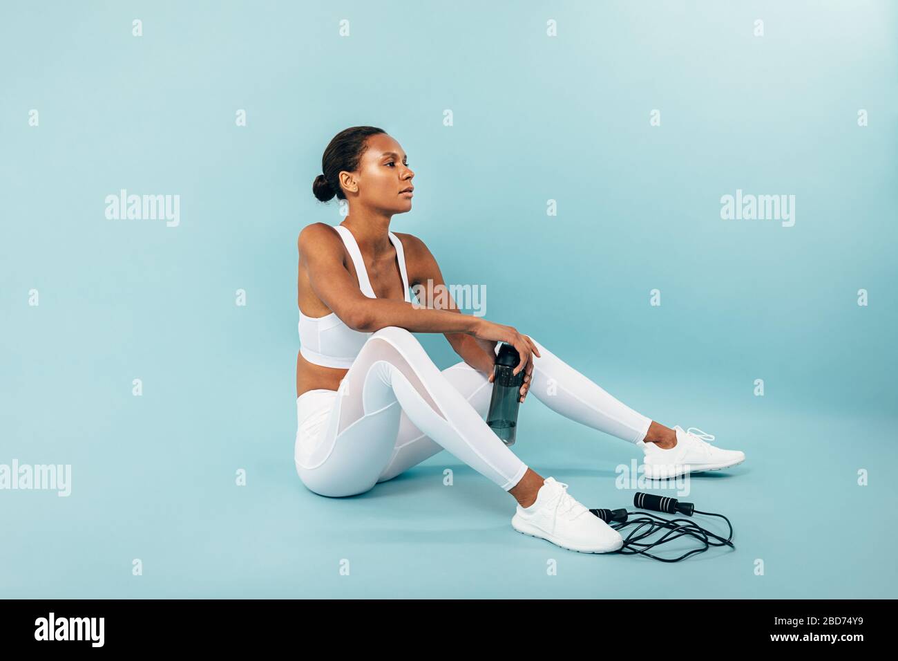 Tired woman in white sportswear sitting on a blue background. Female athlete holding water ...