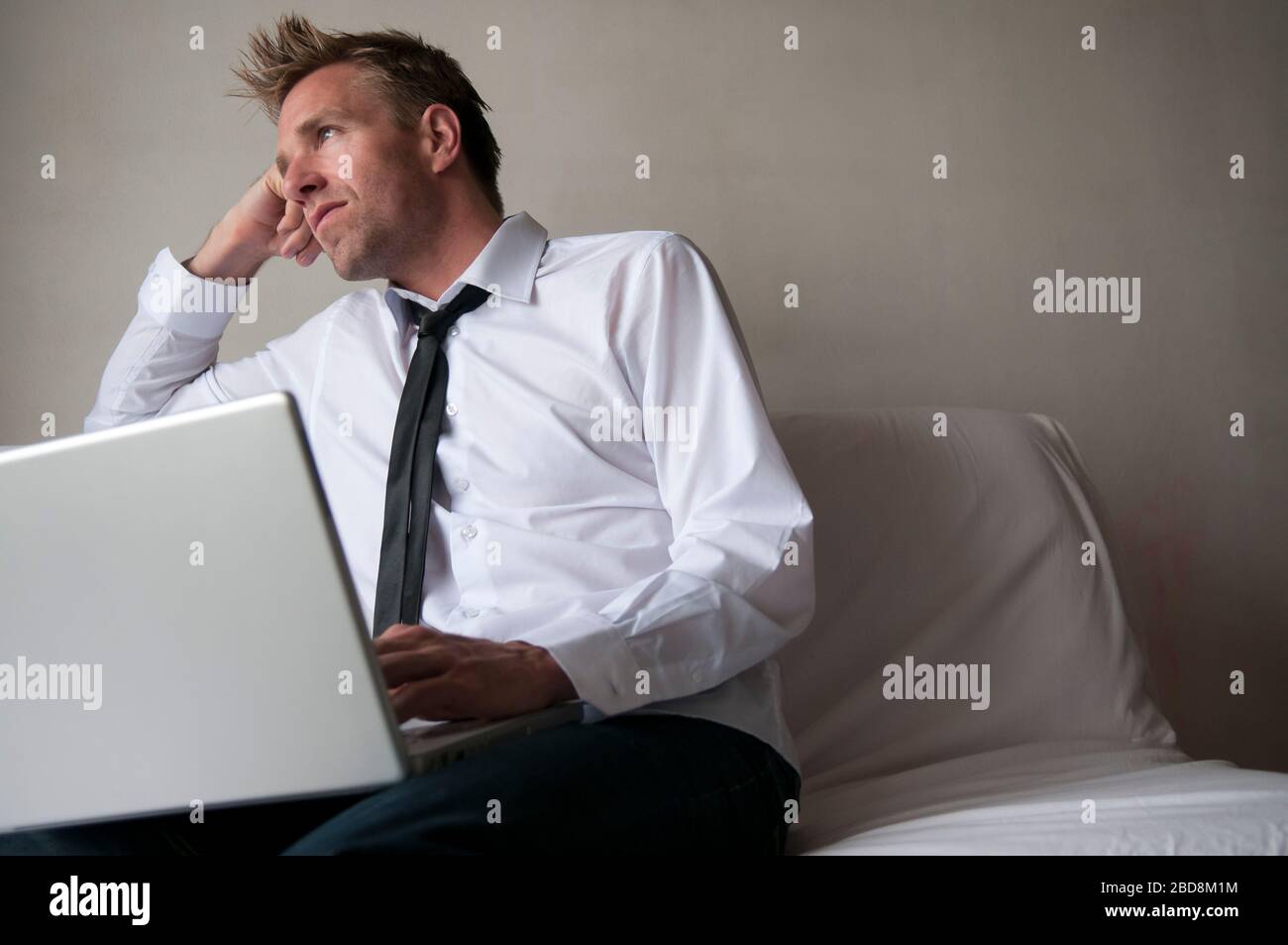 Bored man in smart casual jeans sitting on the sofa browsing his laptop ...