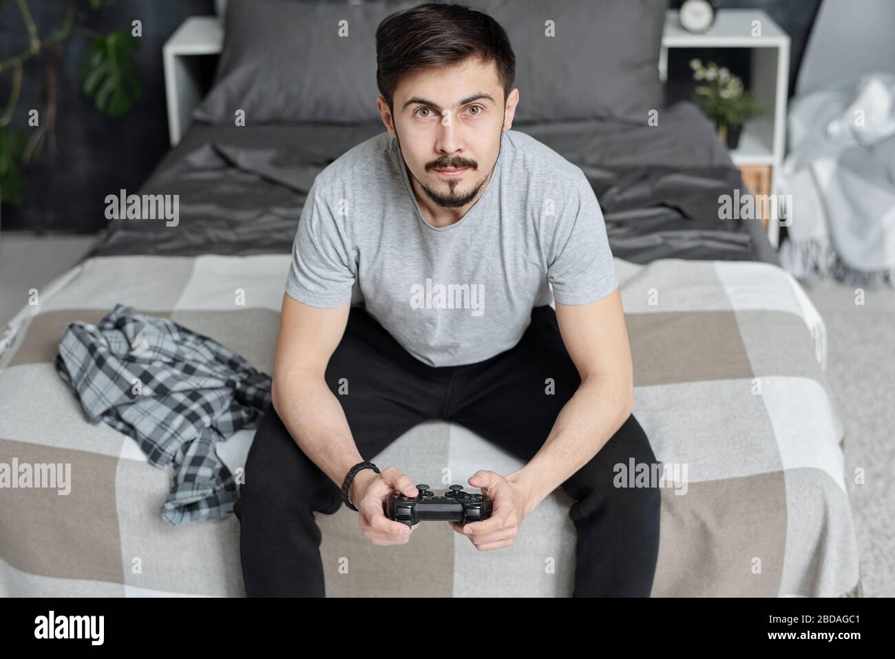 Curious young man sitting on bed and using joystick while playing video ...
