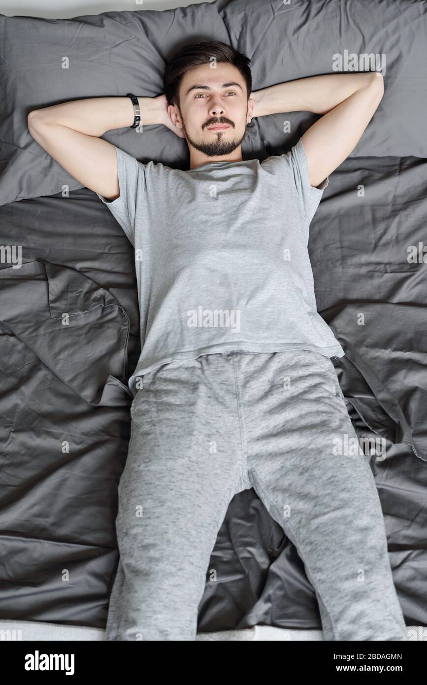 Pensive young bearded man in homewear lying with hands behind head on ...
