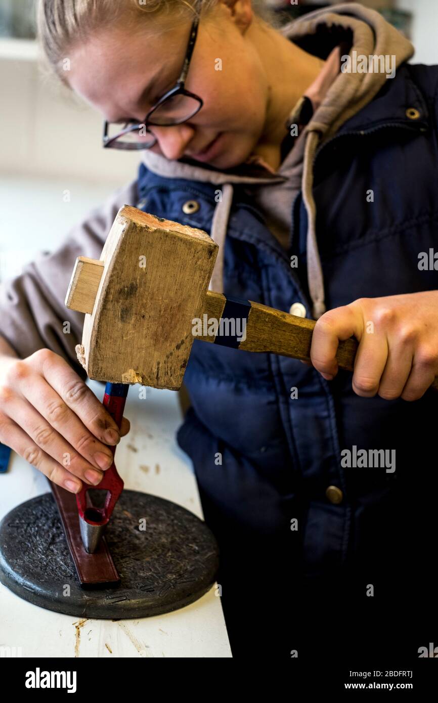 Female saddler standing in workshop, using hand tool and wooden mallet ...