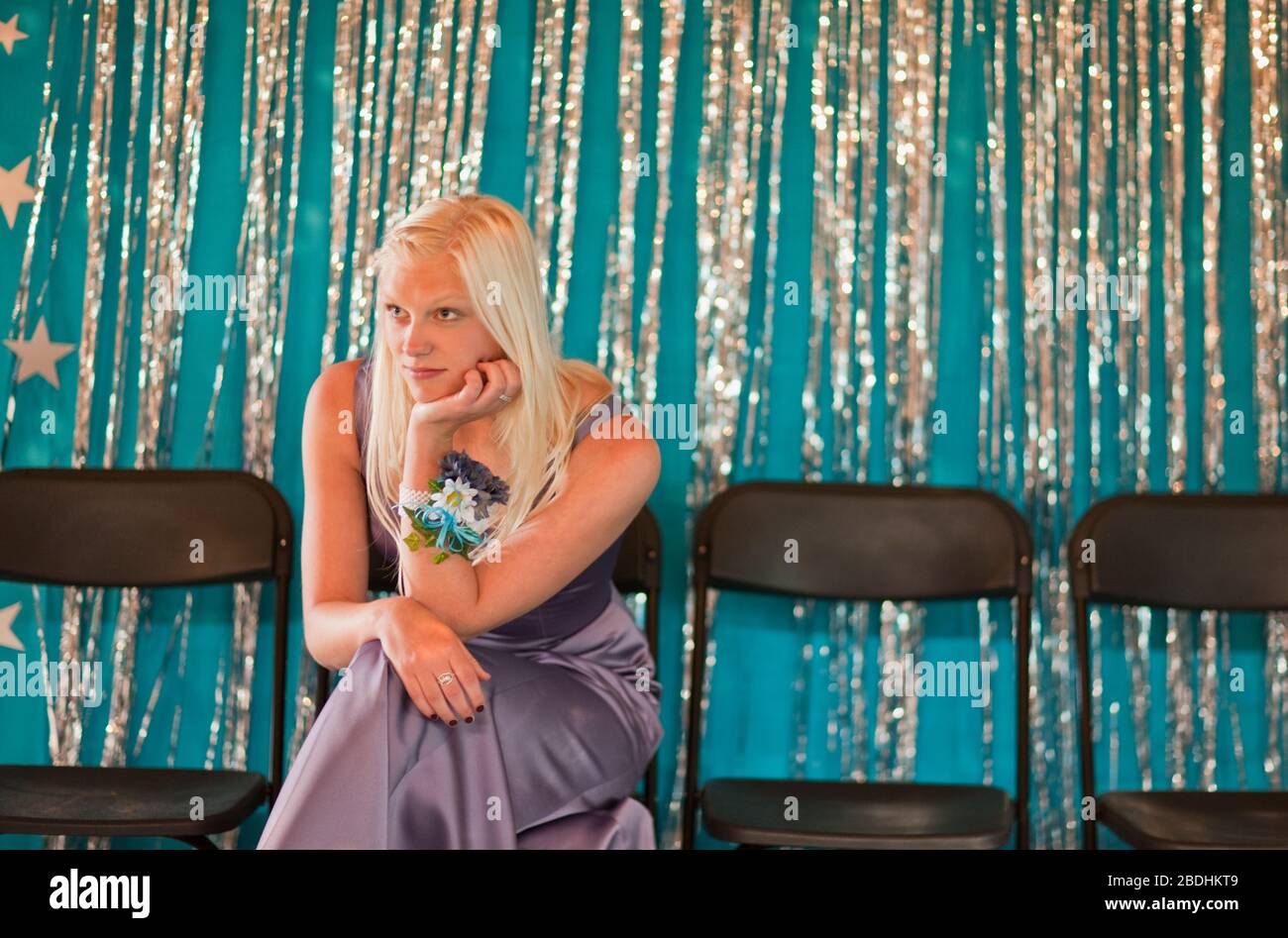 Teenage girl sitting on chair in evening gown, looking bored at prom ...