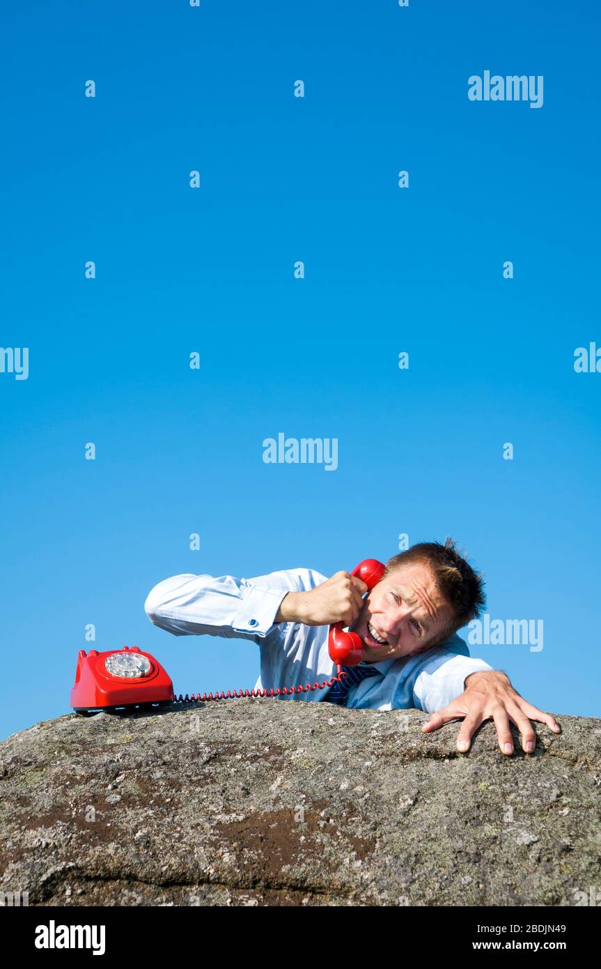 Desperate businessman dangling off a rock cliff making an urgent call ...