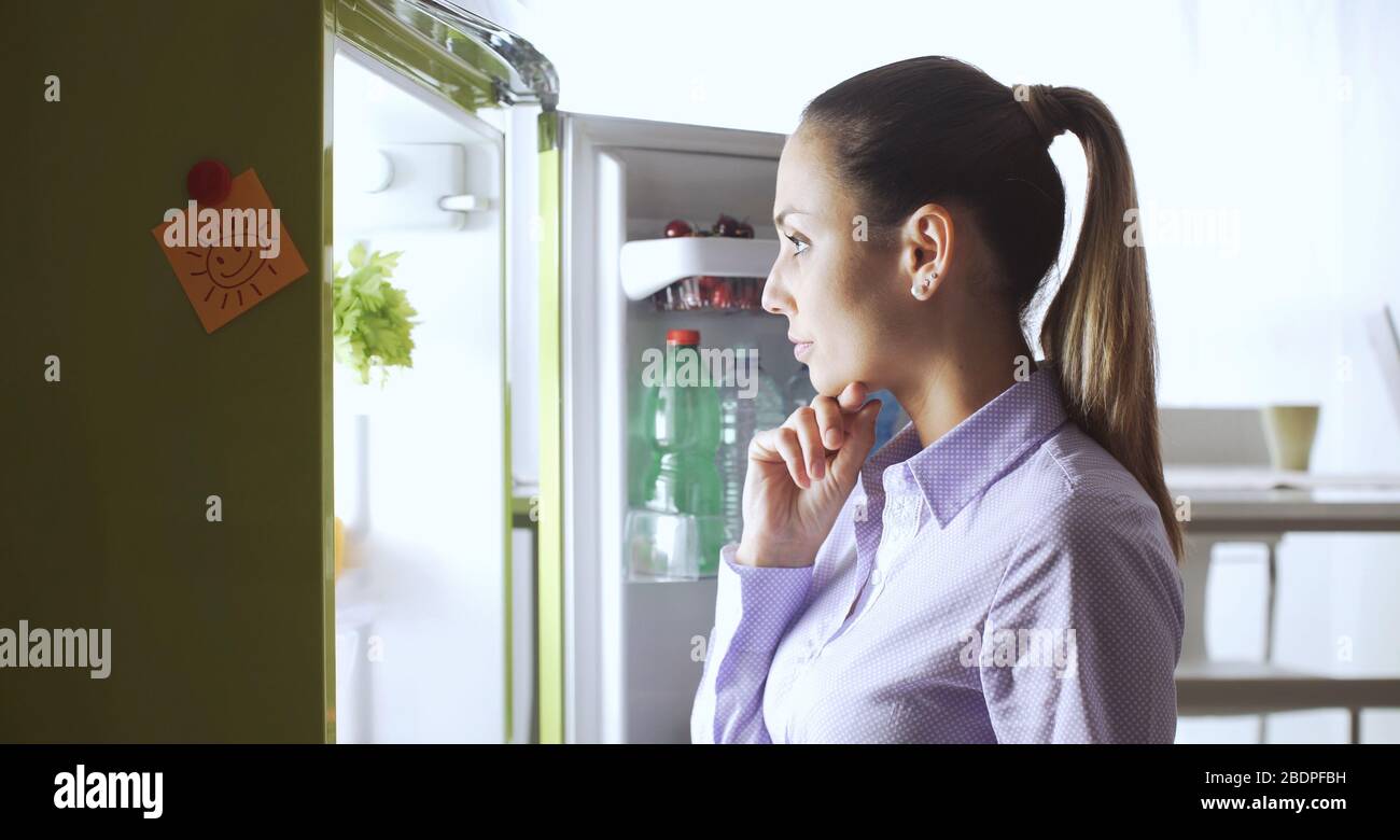 Young woman in the kitchen, she is looking into the fridge and thinking ...