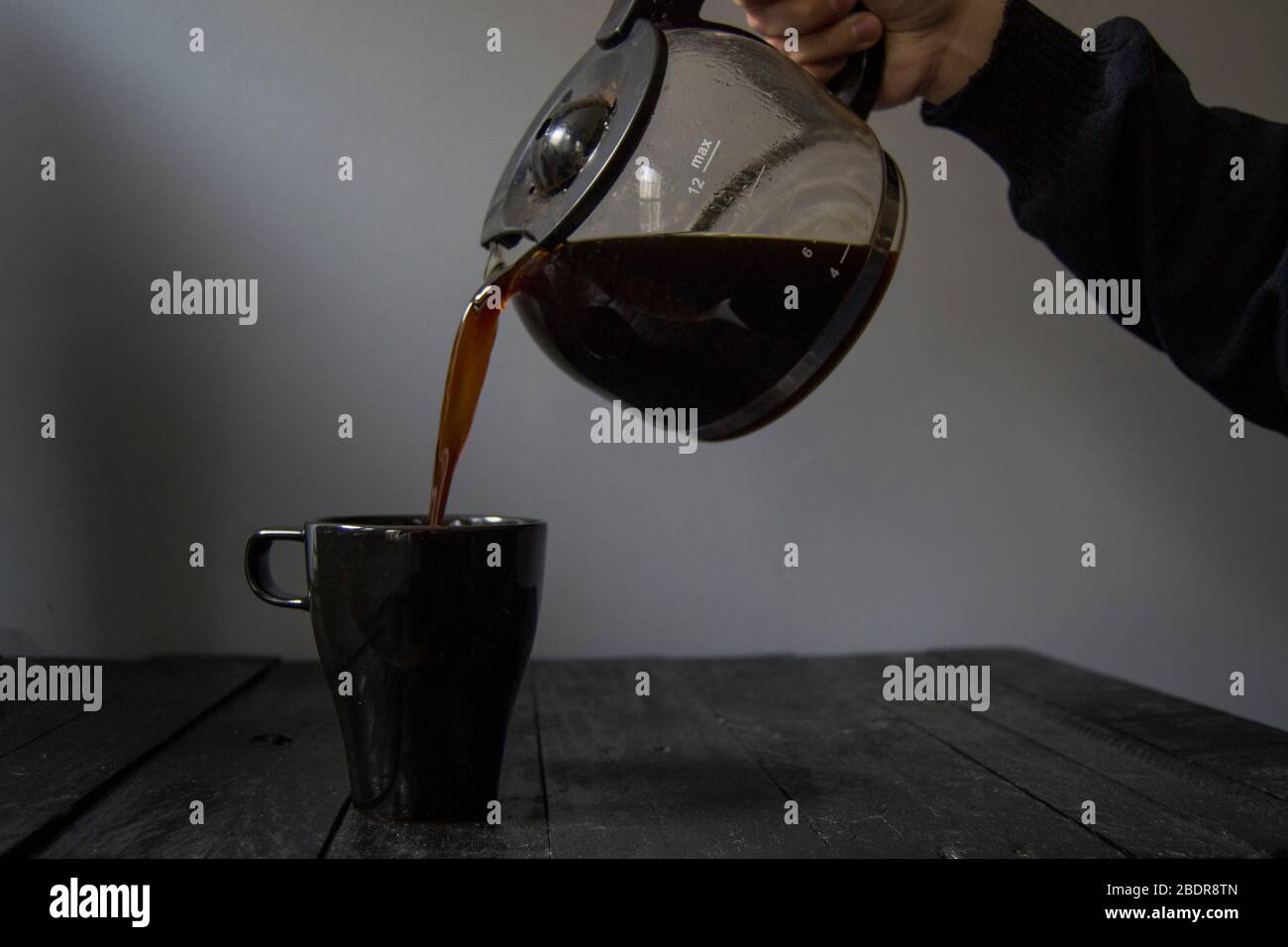 Glass jug pouring coffee into a black mug set on black wood and gray ...