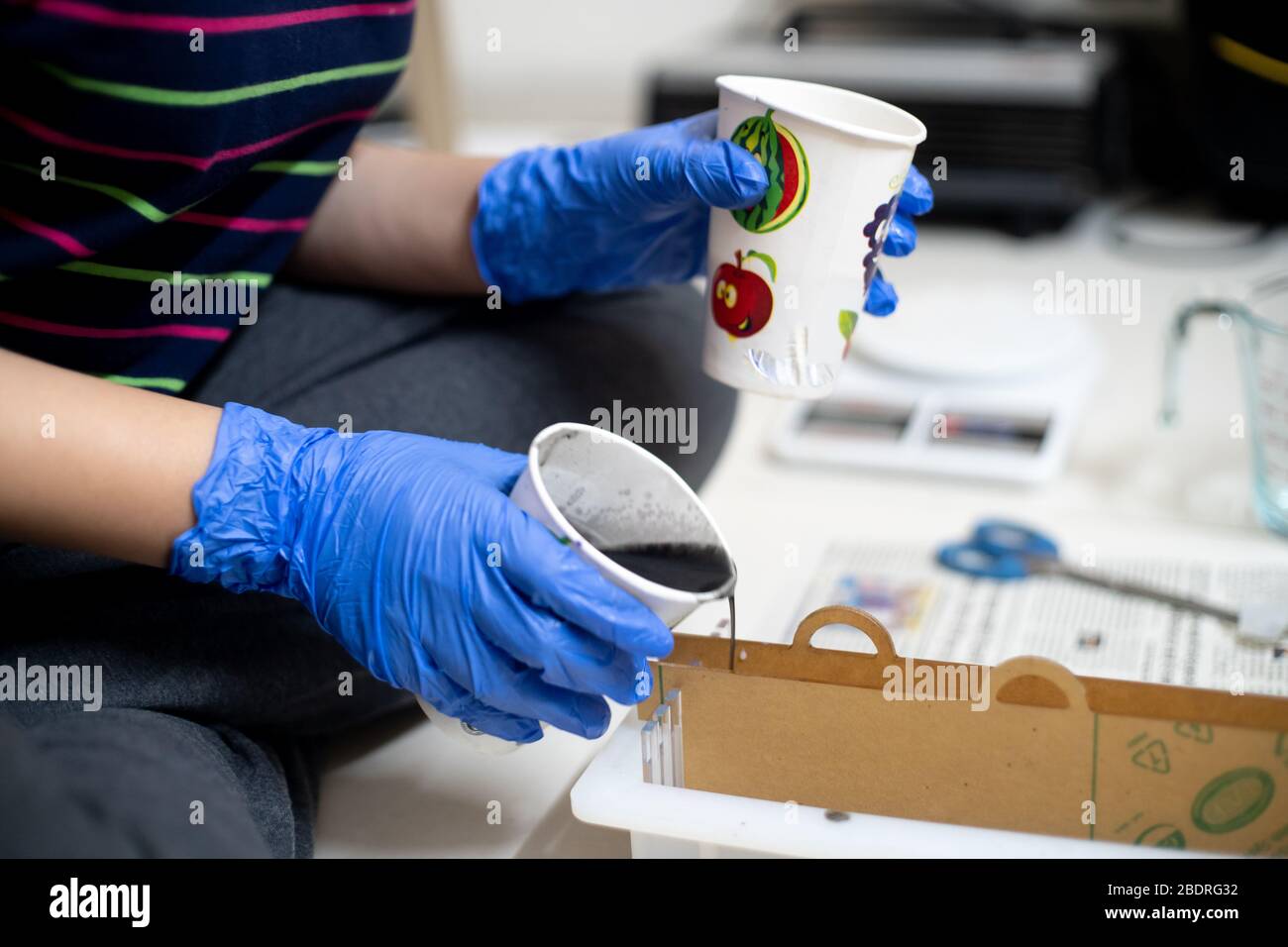 Blue gloved hand pouring red liquid from a paper cup into a mold with ...