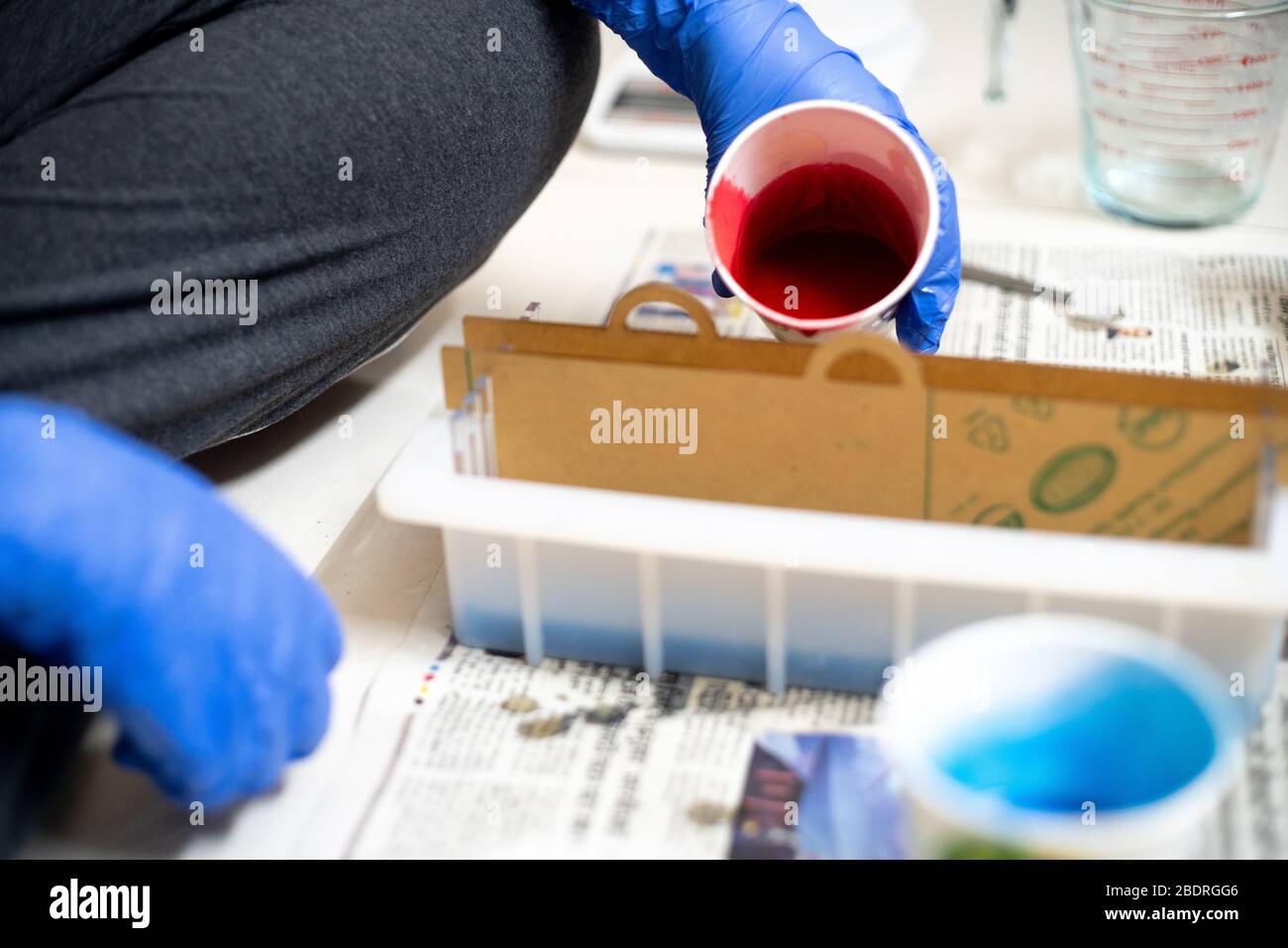Blue gloved hand pouring red liquid from a paper cup into a mold with ...