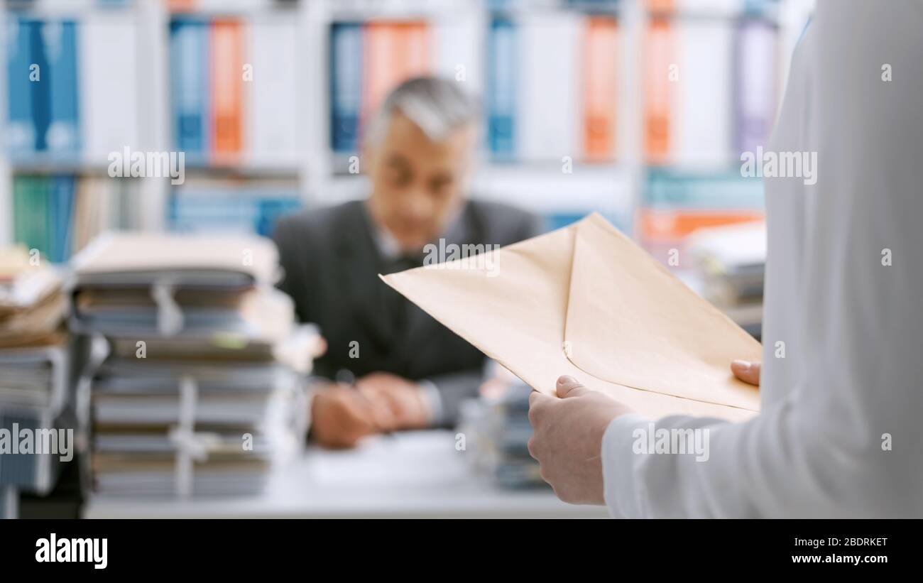 Assistant bringing mail to her boss, he is working at desk overloaded ...