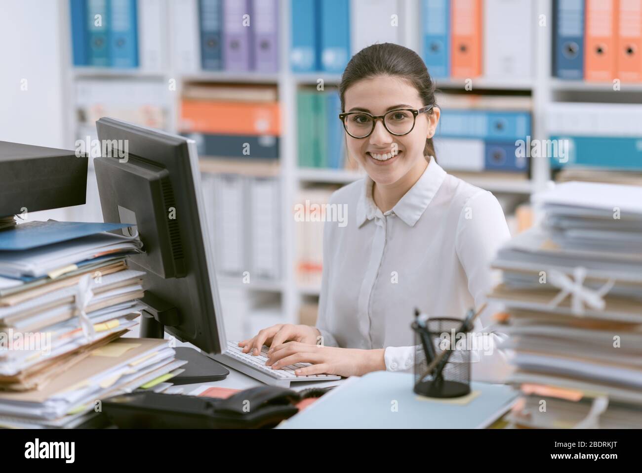 Young smiling secretary working at office desk and stacks of paperwork ...