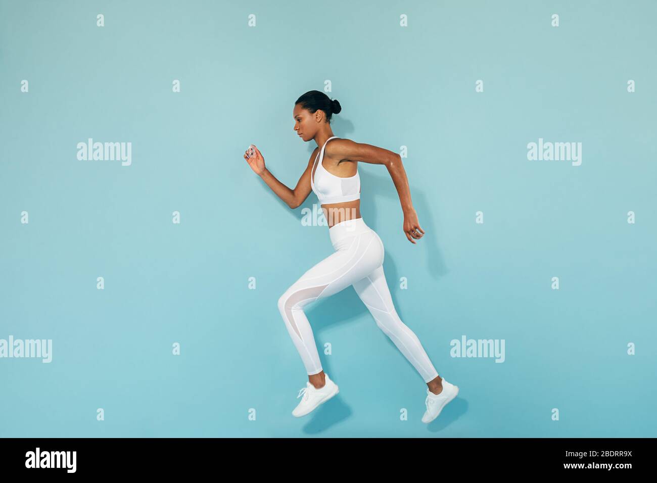 Side view of a woman running and jumping over blue background in studio ...