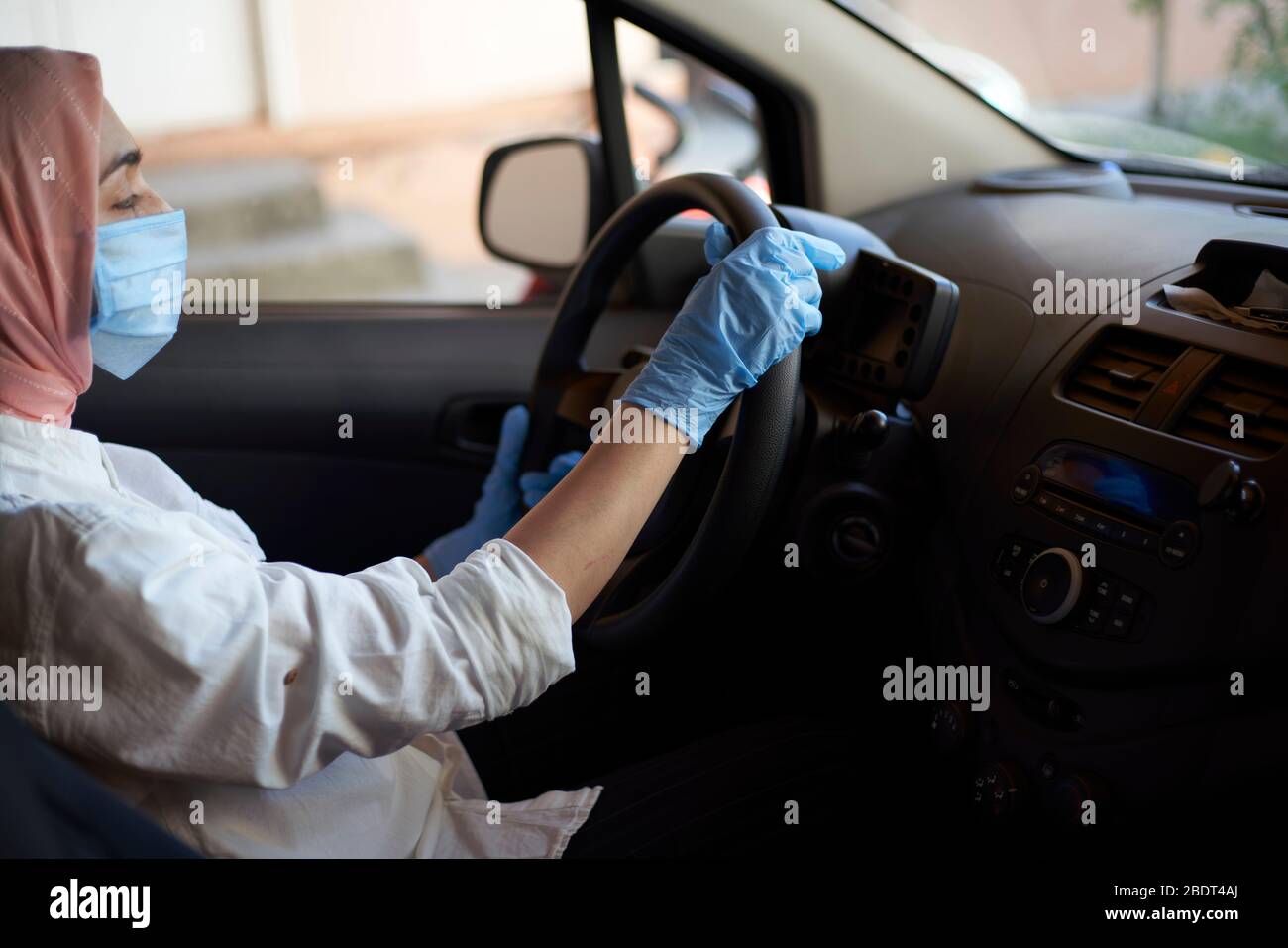 A young Muslim woman in a surgical face mask and gloves driving the car ...