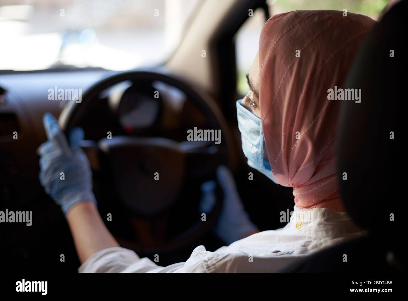 A young Muslim woman in a surgical face mask and gloves driving the car ...
