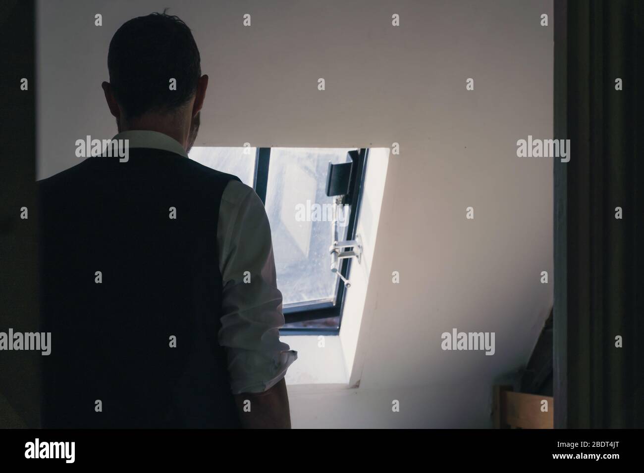 A man looking out of a window, back to camera, wearing a waistcoat ...