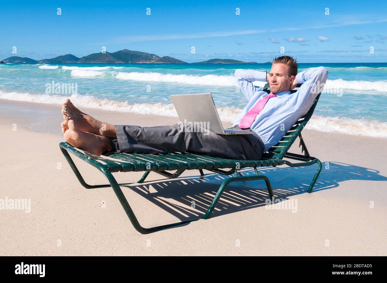 Barefoot businessman working on his laptop on a beach chair relaxing on ...