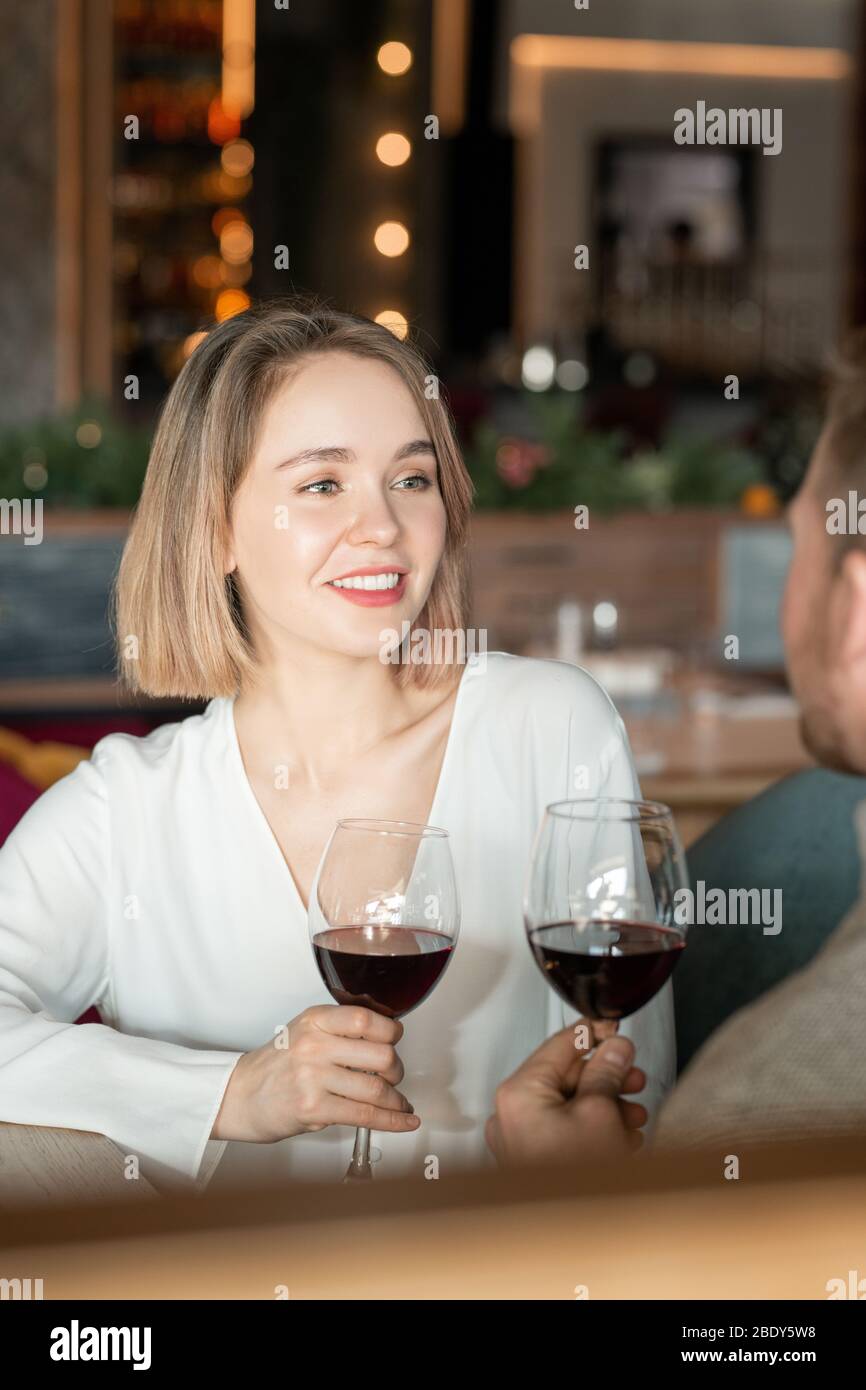 Pretty young woman with toothy smile talking to her boyfriend while making toast with glasses of ...