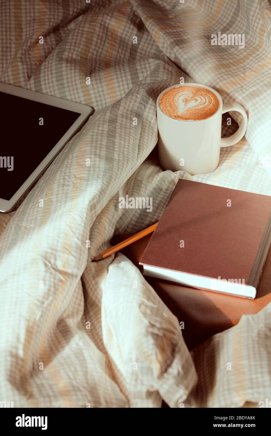 Notepad, book, laptop and cup of coffee on checkered textile in bed ...