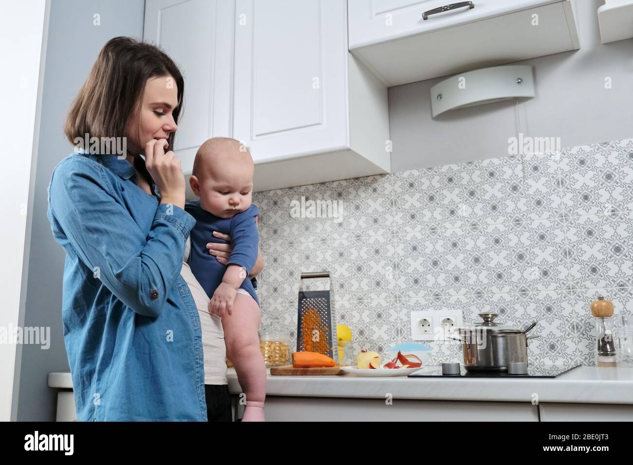 Young beautiful woman with baby in her arms in kitchen. Mother with son cooking food in pan in ...