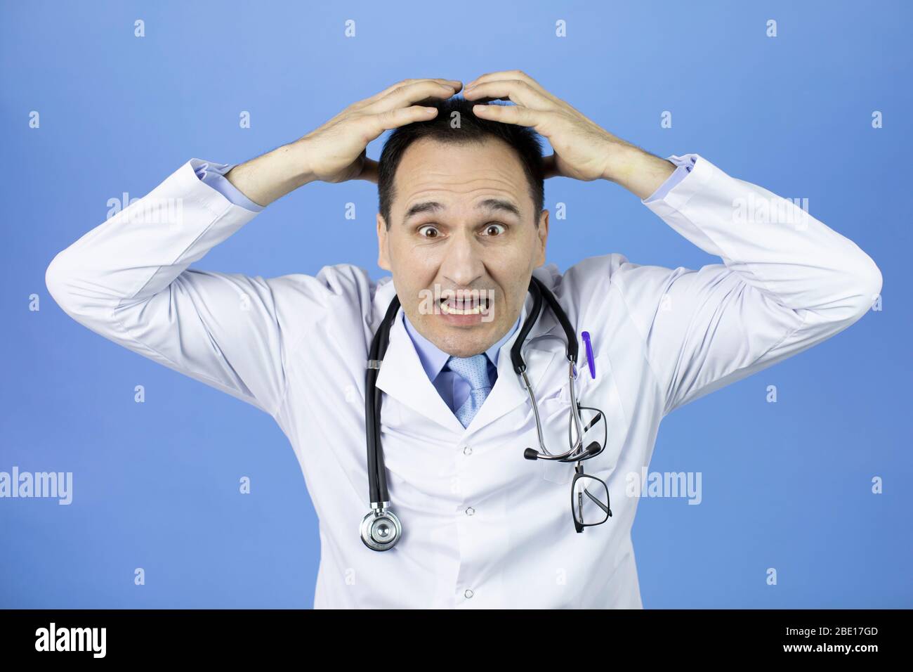 Shocked doctor shouting with fear with raised hand on head over blue ...