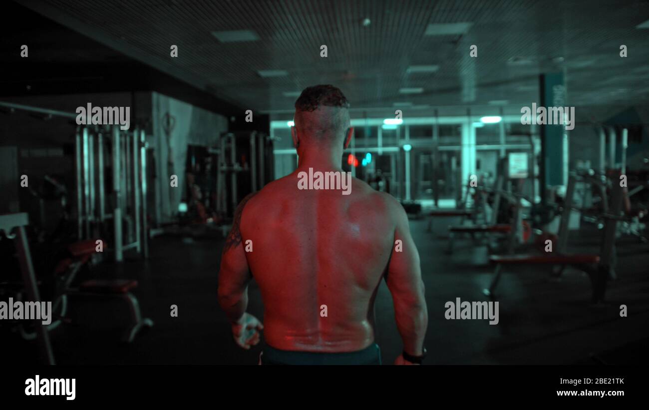 An adult shirtless man bodybuilder walking in the dark gym with neon ...