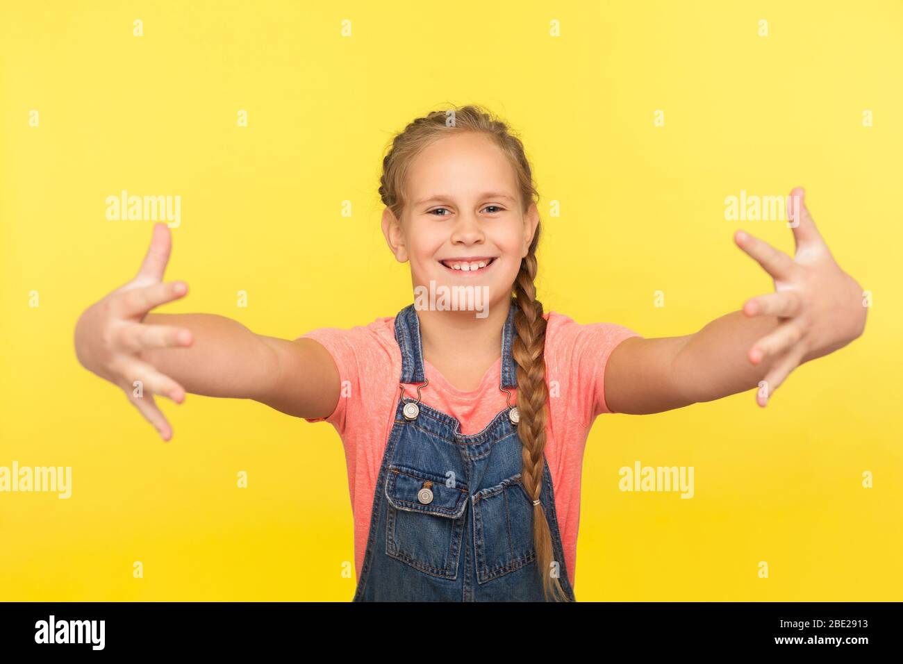 Portrait of excited generous little girl with braid in denim overalls ...