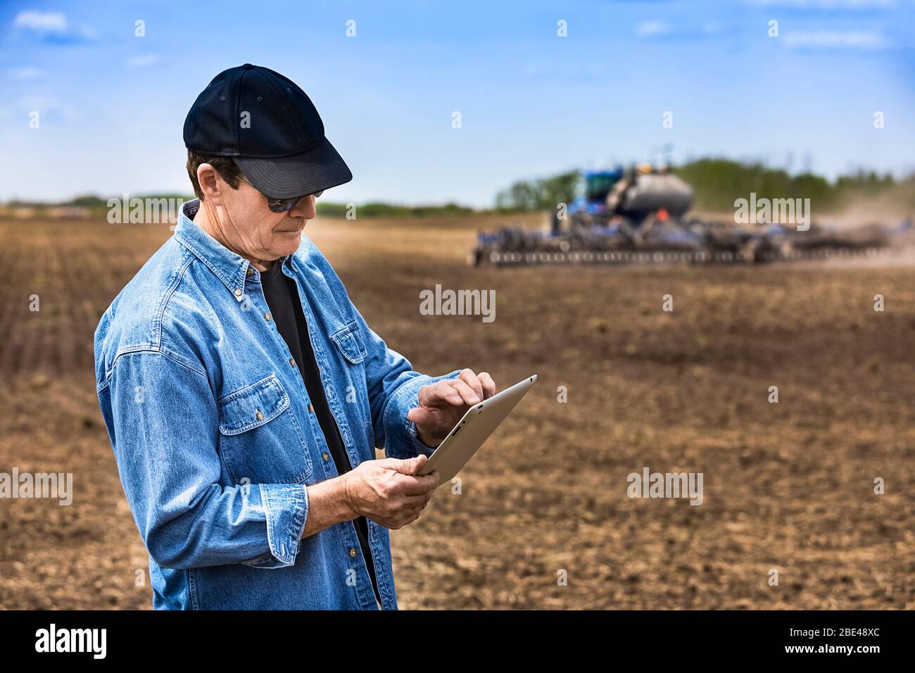 Farmer using a tablet while standing on a farm field and a tractor and ...