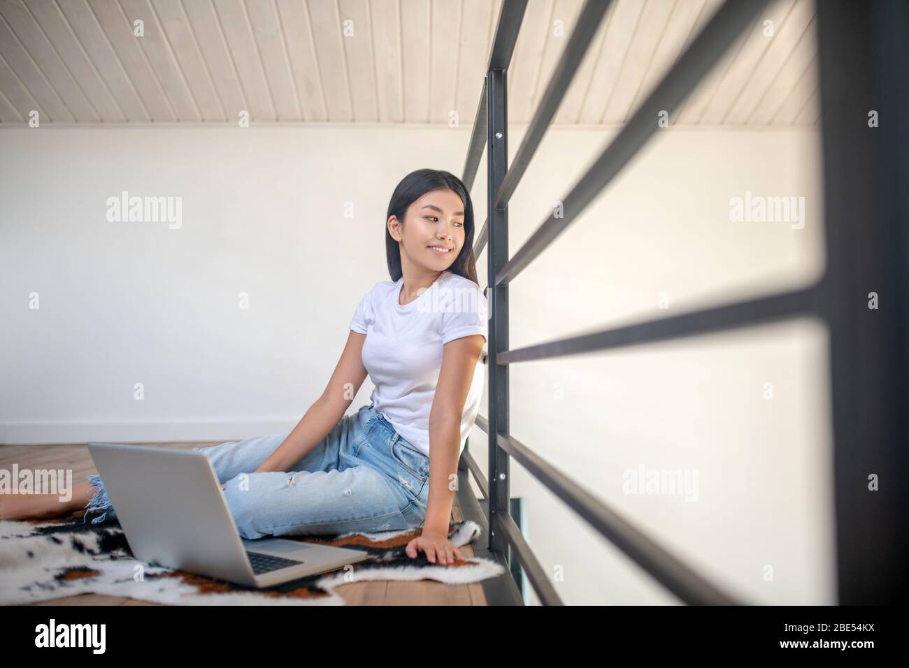 Young woman sitting on the floor with laptop, leaning on rails, looking ...