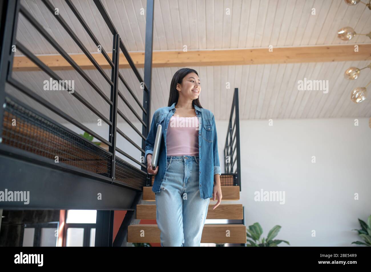 Young woman going downstairs, holding laptop under arm, looking ...