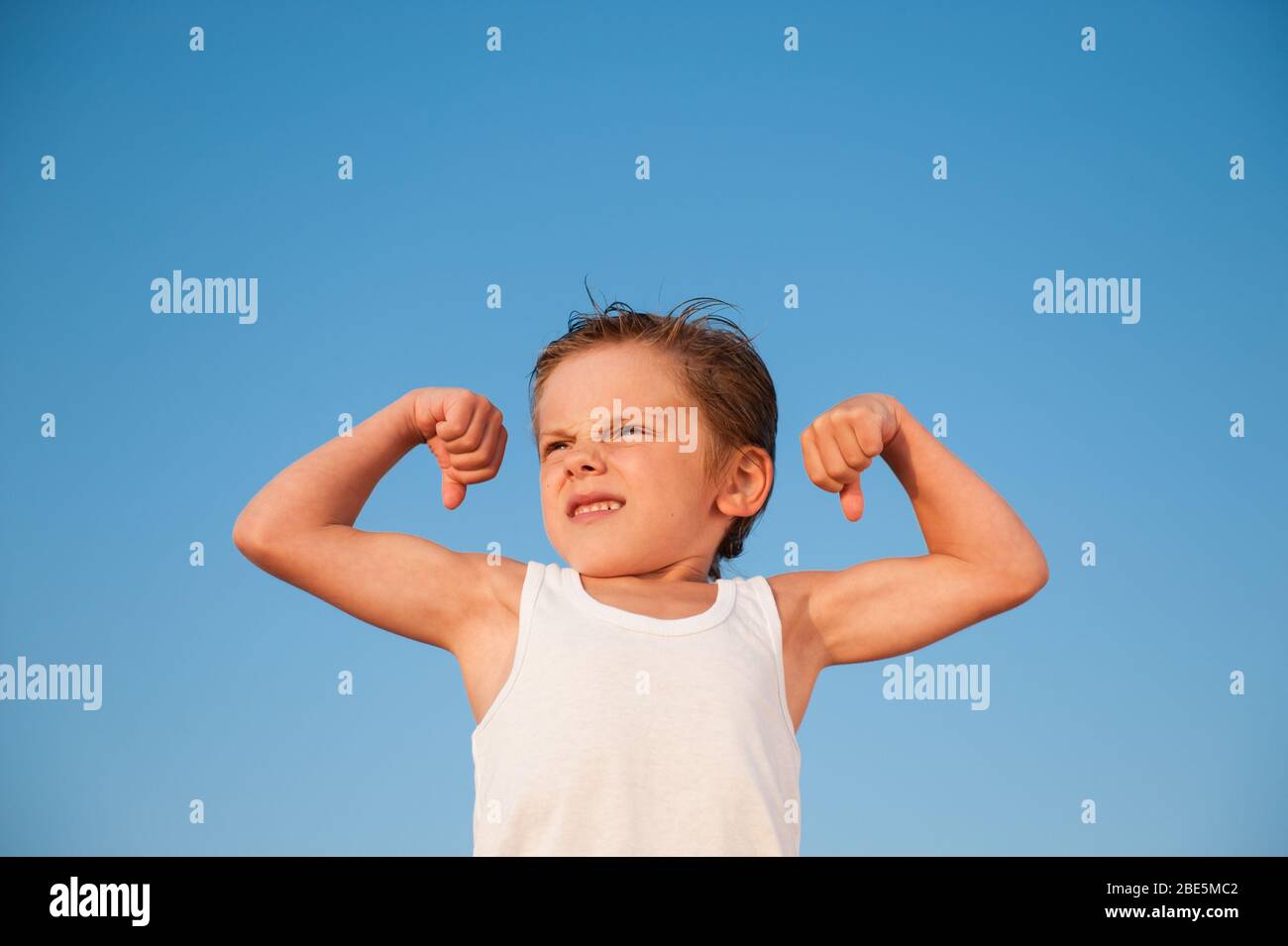 strong little boy in white tank top showing strong muscle on blue sky ...