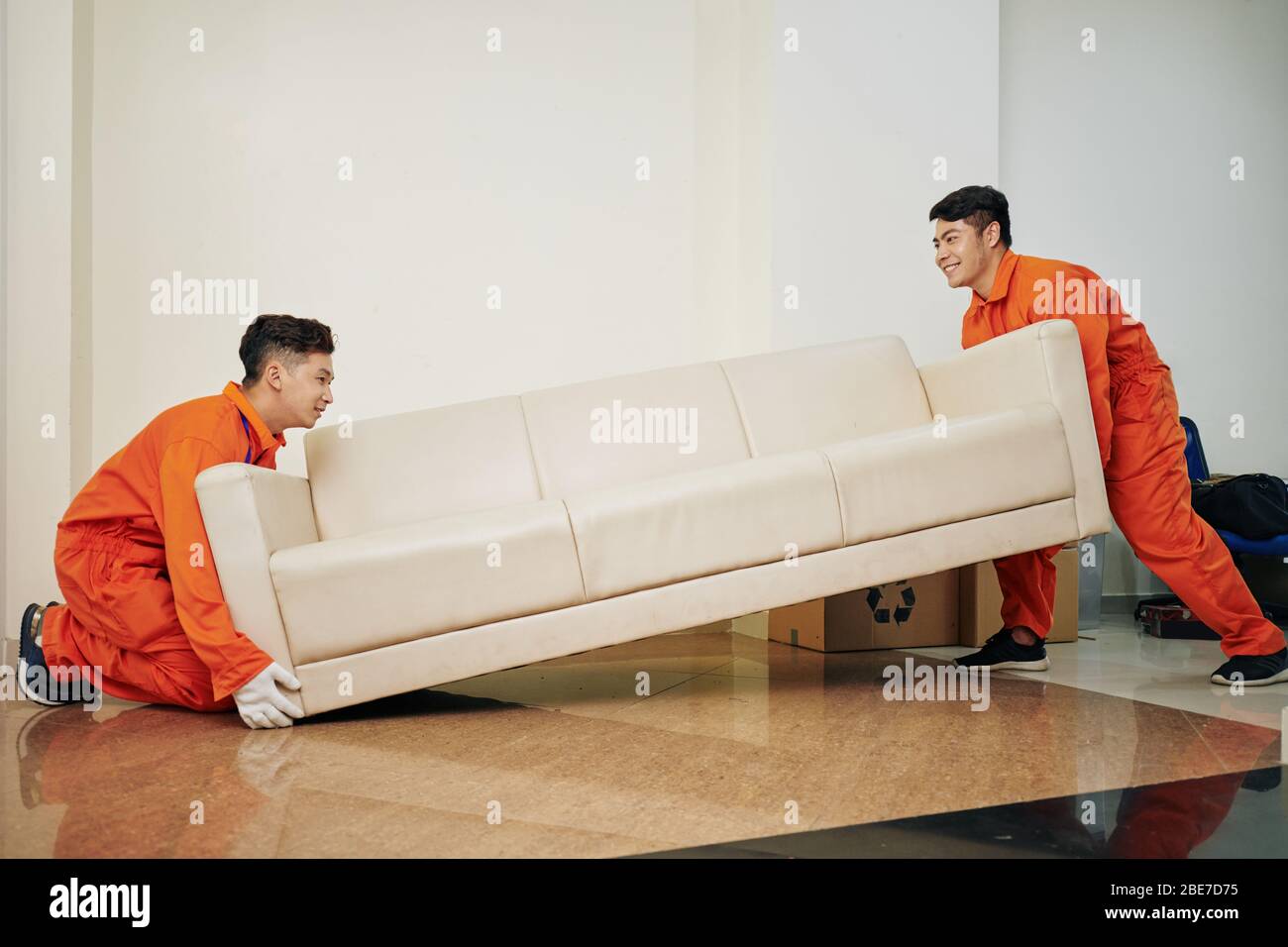 Smiling moving service company workers in uniform carrying heavy couch ...
