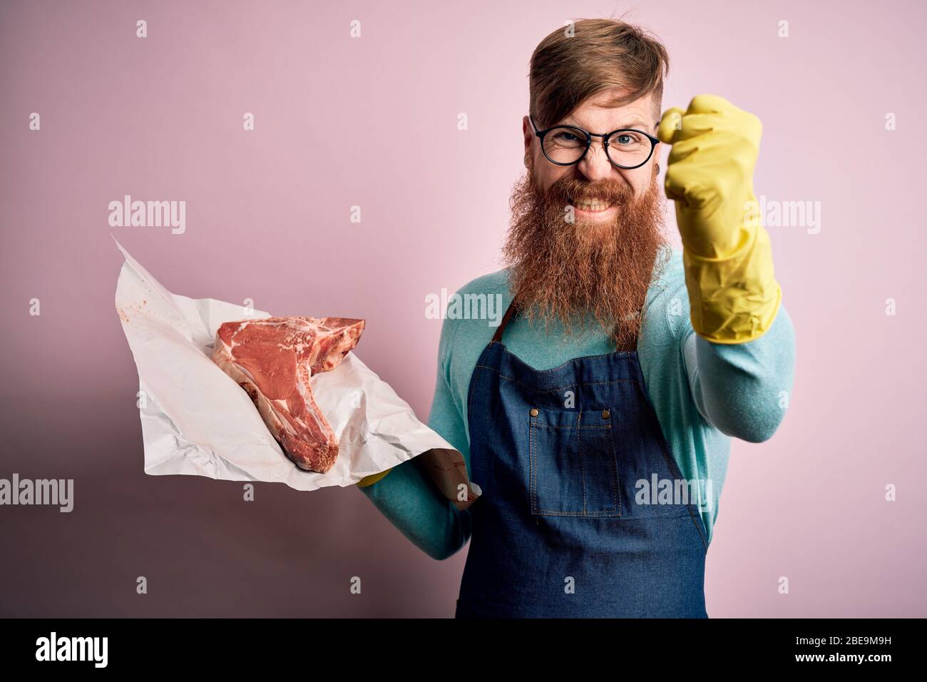 Redhead Irish butcher man with beard holding raw beef steak over pink ...