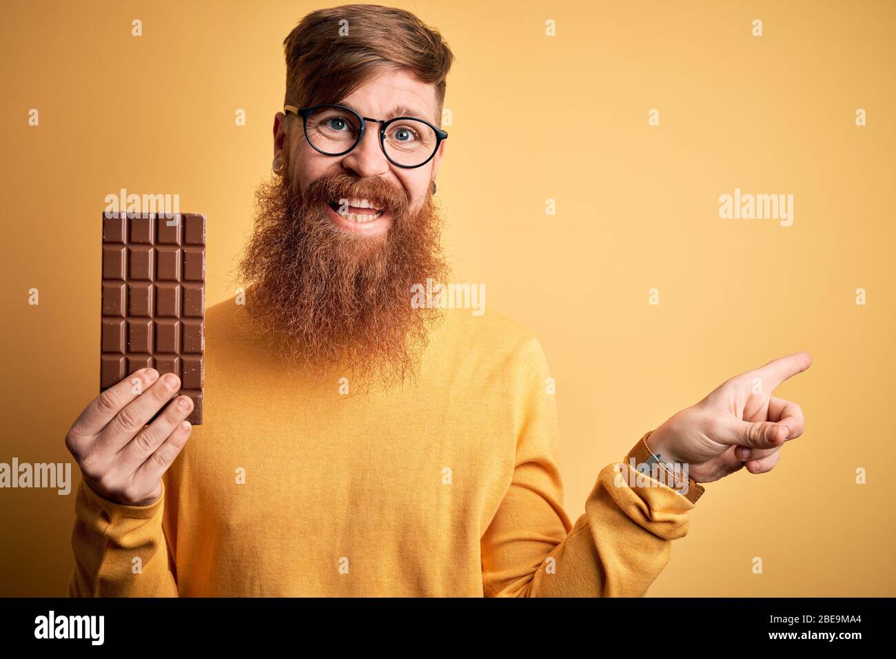 Irish redhead man with beard eating a sweet chocolate bar over yellow ...