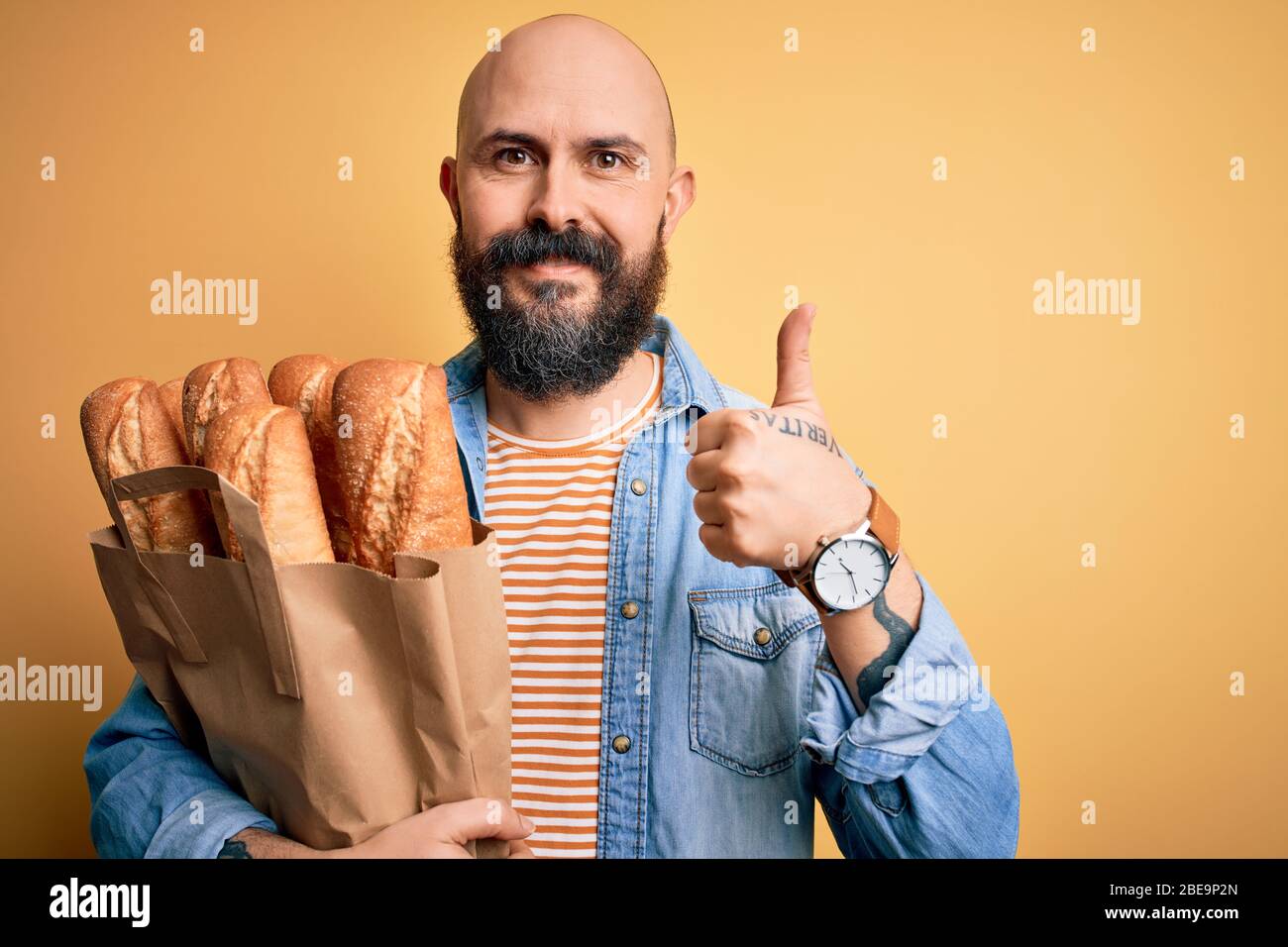 Handsome bald man with beard holding paper bag with bread over yellow background happy with big ...