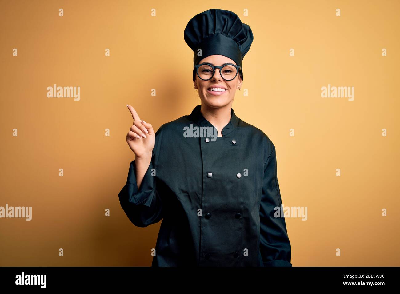 Young beautiful brunette chef woman wearing cooker uniform and hat over ...
