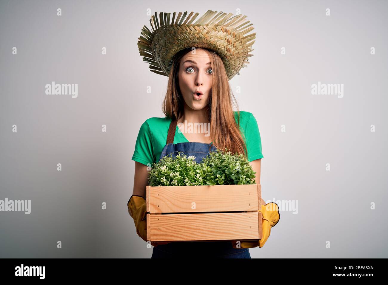 Young beautiful redhead farmer woman wearing apron and hat holding box ...
