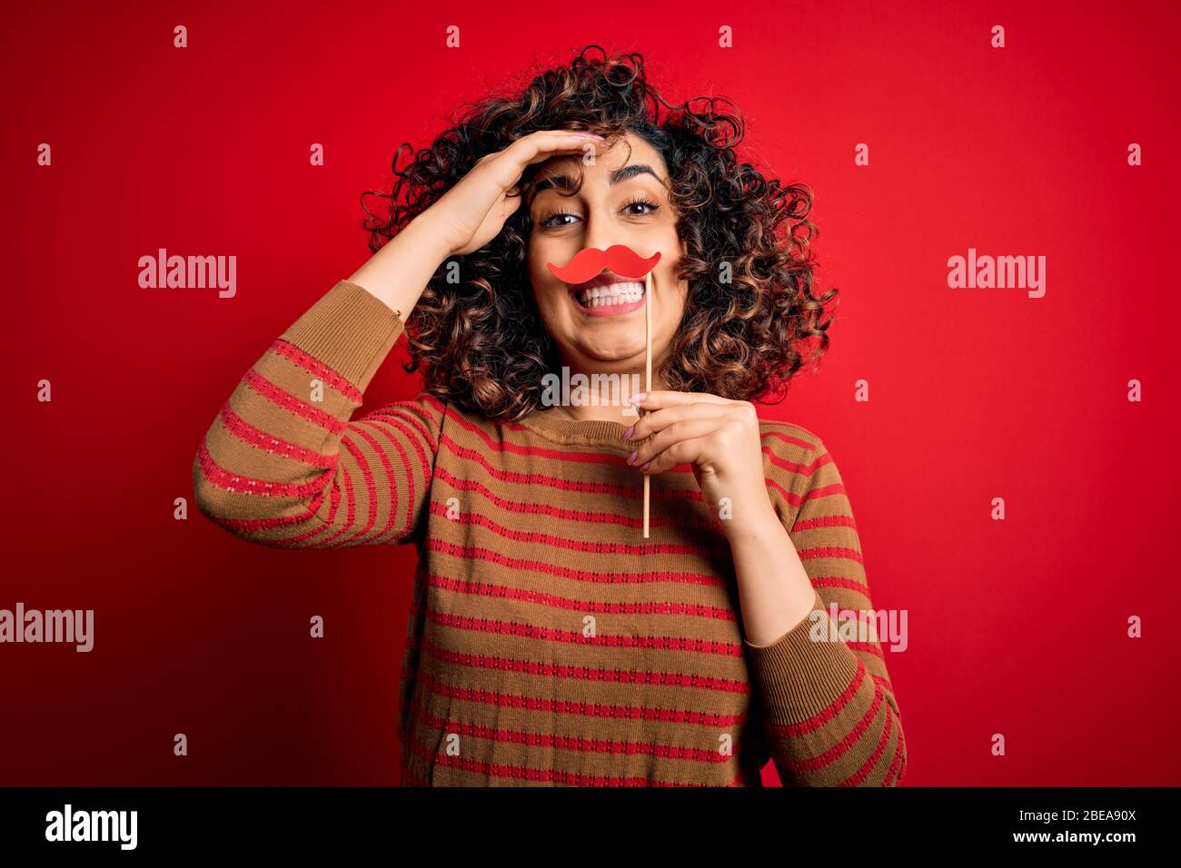 Young beautiful curly arab woman holding funny mustache over isolated ...