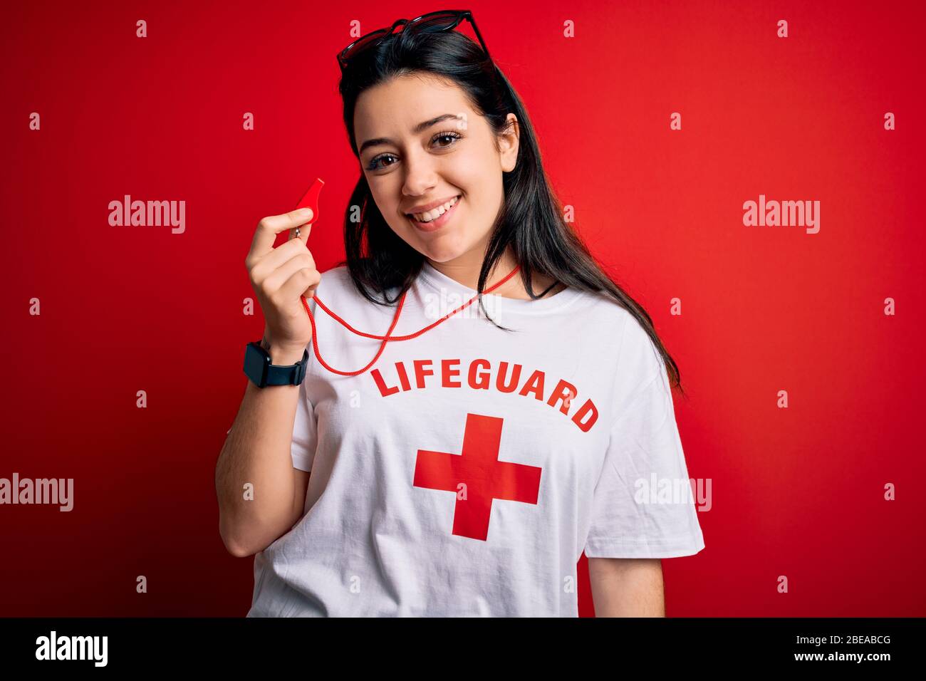 Young lifeguard woman wearing guard equipement holding whistle over red ...
