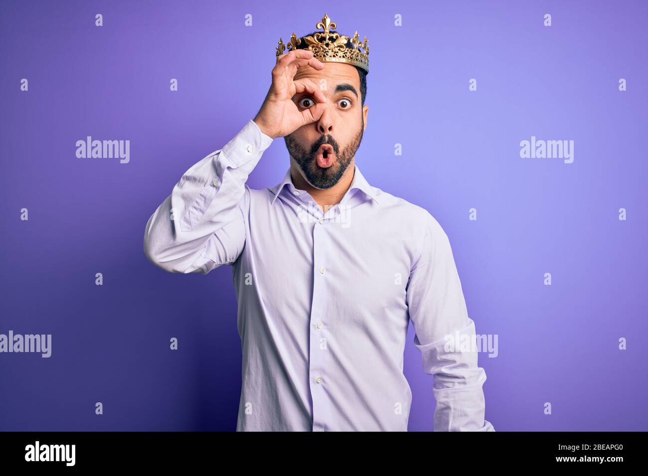 Young handsome man with beard wearing golden crown of king over purple ...
