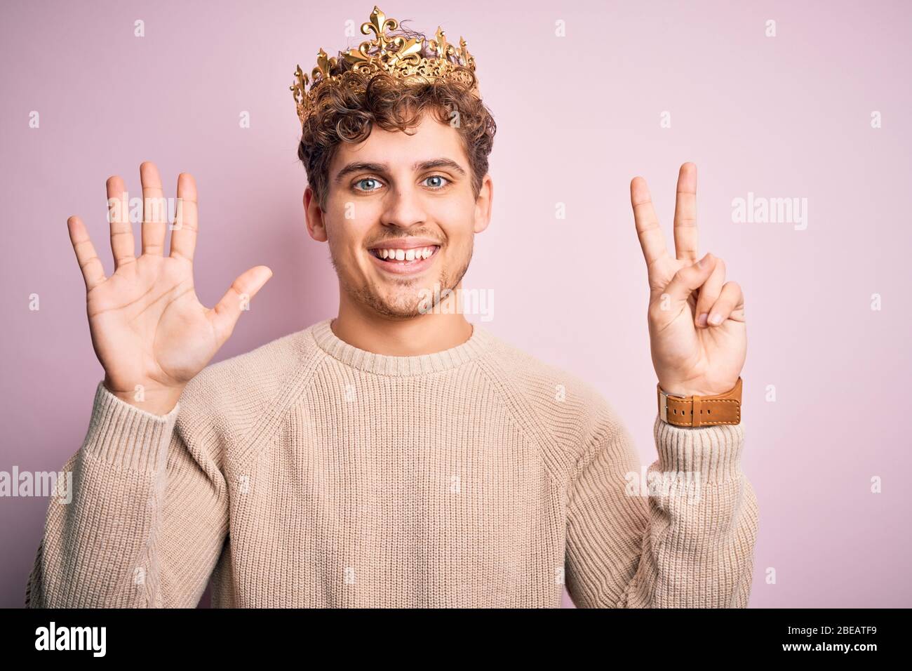 Young blond man with curly hair wearing golden crown of king over pink ...