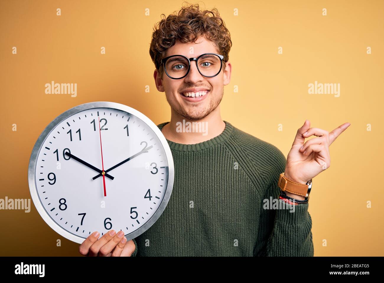 Young blond man with curly hair wearing glasses holding big clock over ...