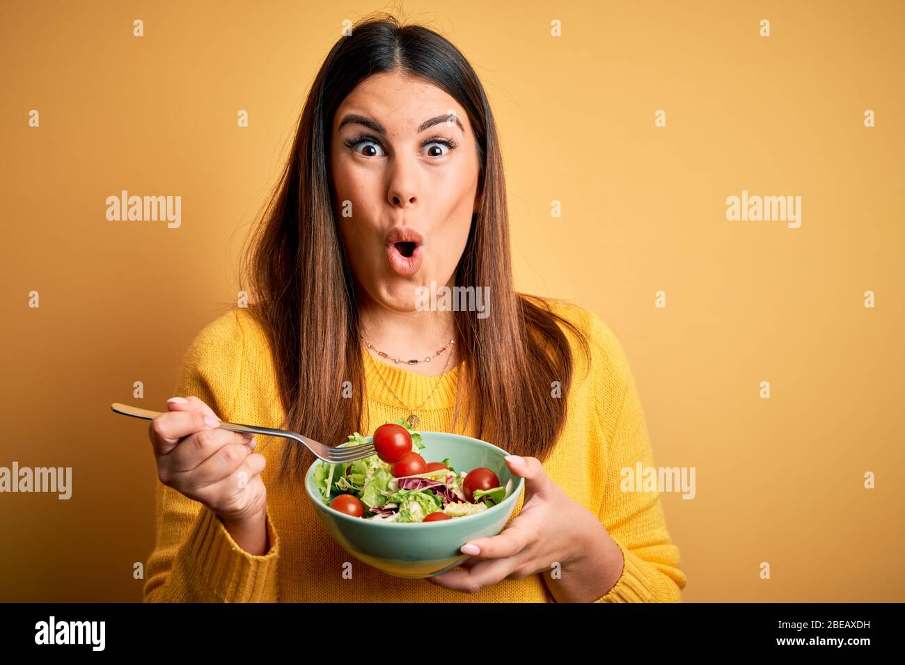 Young beautiful woman eating healthy fresh salad over yellow background ...