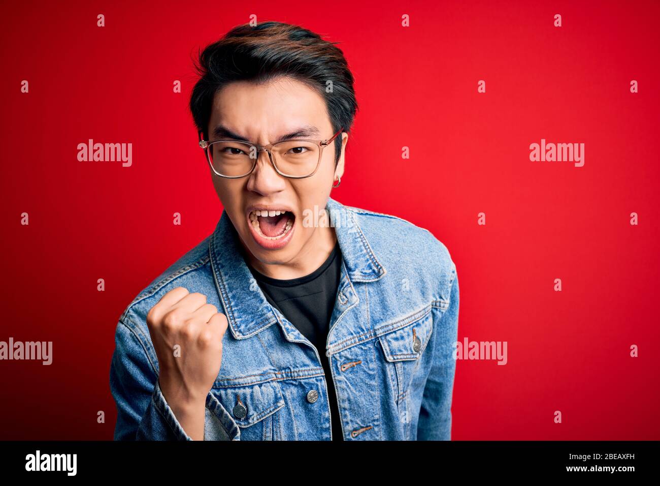 Young handsome chinese man wearing denim jacket and glasses over red ...
