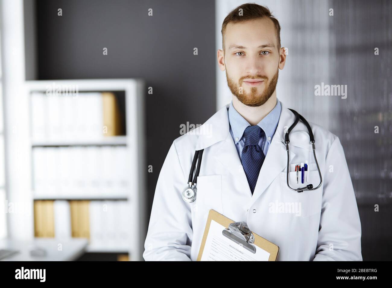 Red-bearded doctor standing straight in clinic near his working place ...