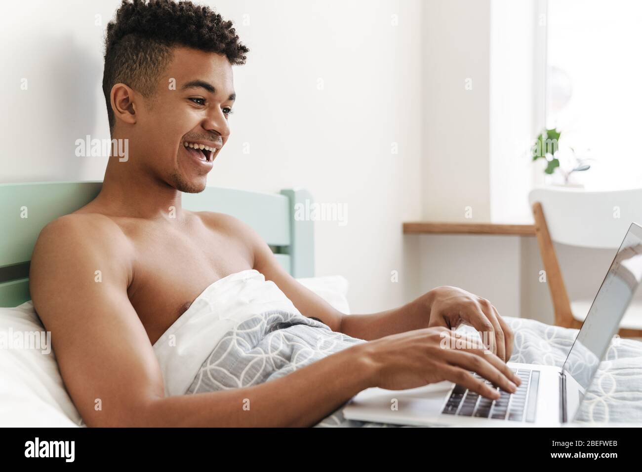 Image of young happy african guy in the morning in bedroom using laptop ...
